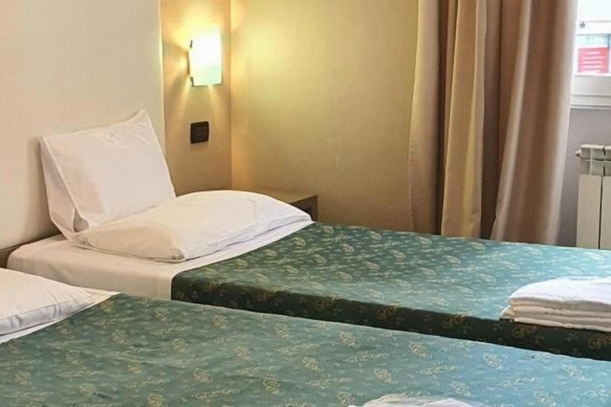 Quarto de hotel com duas camas, uma luminária fixada na parede e uma janela com cortinas.