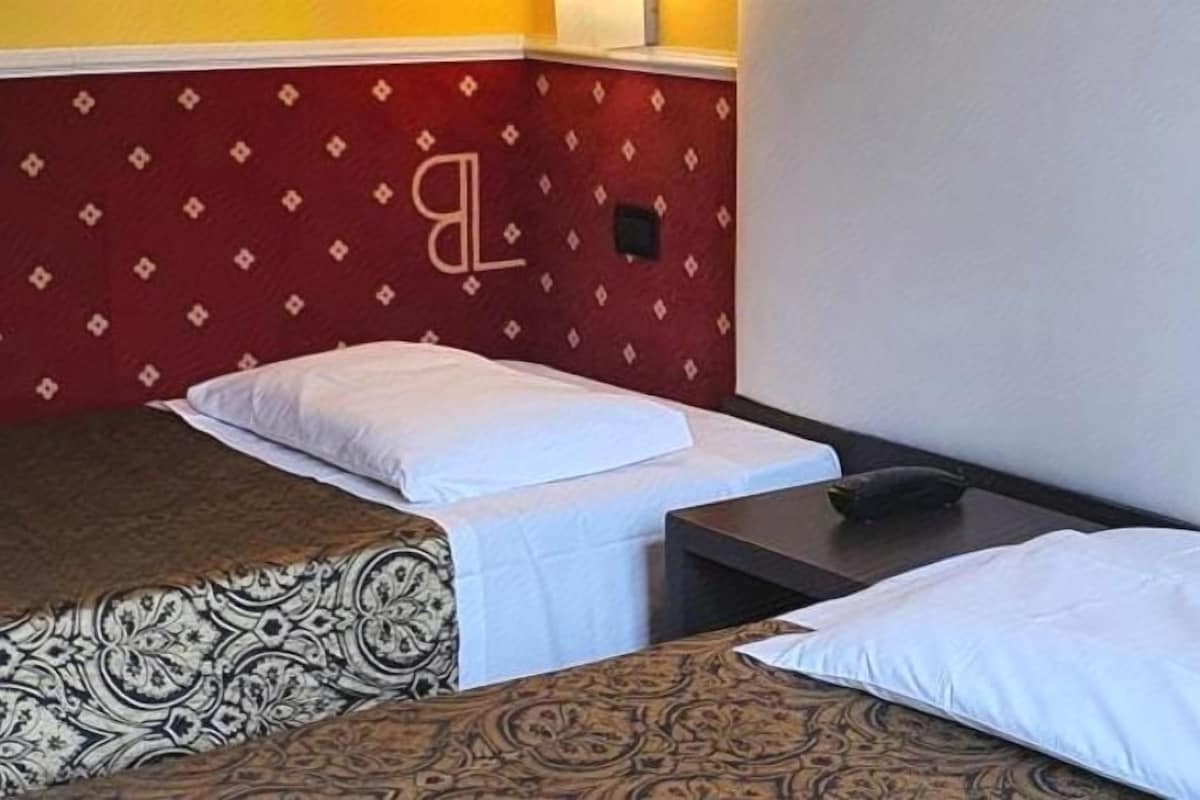 Quarto de hotel com duas camas, cada uma com roupa de cama estampada, uma pequena mesa de cabeceira e uma luminária fixada na parede.