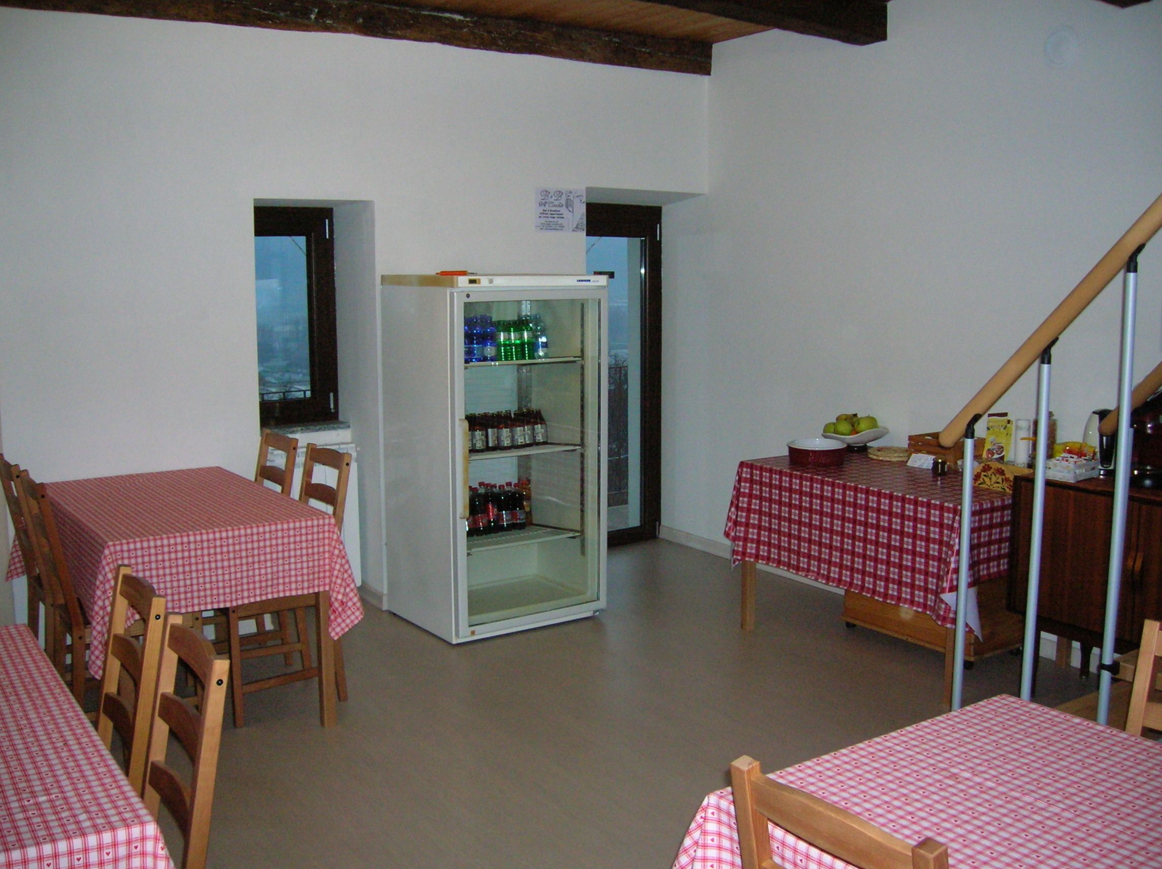 Uma área de refeições com toalhas de mesa xadrez vermelhas e brancas, cadeiras de madeira, um refrigerador cheio de bebidas e uma janela com vista.