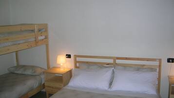 Uma cama de solteiro e uma cama de casal em beliche, com estrutura de madeira. Há uma mesa de cabeceira com abajur e uma tomada na parede.