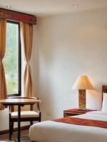Quarto de hotel com cama, mesa, uma cadeira, luminária e janela com cortinas.