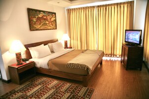 Quarto de hotel com uma cama, duas luminárias de cabeceira, uma televisão sobre um suporte de madeira, um tapete estampado e um quadro na parede.