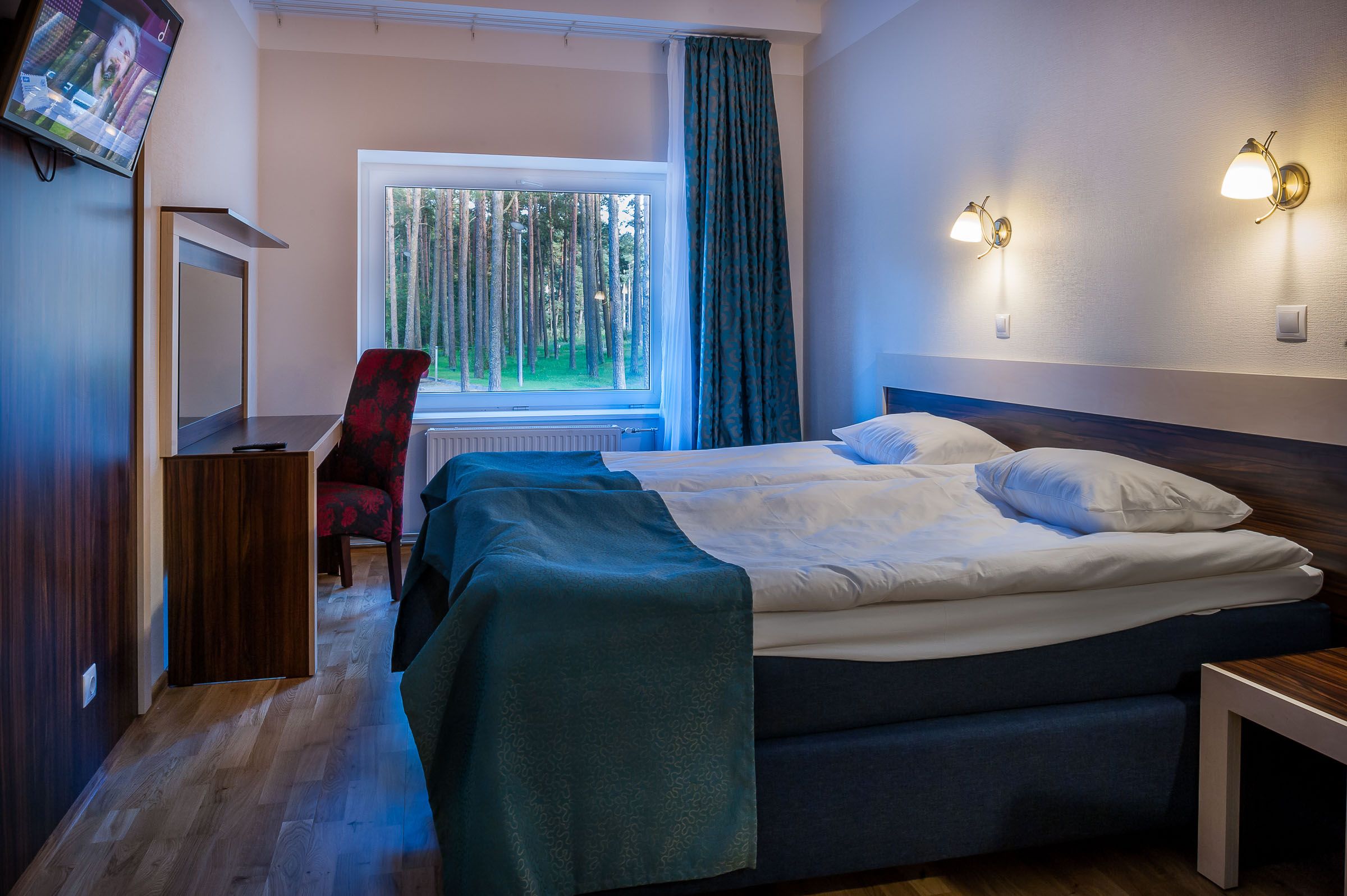 Quarto de hotel com cama, escrivaninha, cadeira, televisão e janela com cortinas.