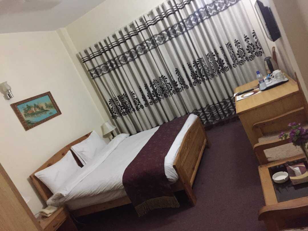Quarto de hotel com uma cama de solteiro, uma mesa de cabeceira de madeira, uma escrivaninha com cadeira e cortina estampada com padrão floral.