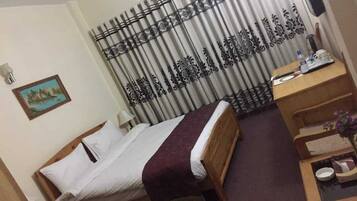 Quarto de hotel com uma cama de solteiro, uma mesa de cabeceira de madeira, uma escrivaninha com cadeira e cortina estampada com padrão floral.