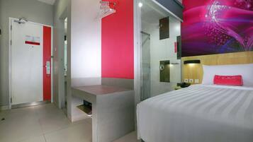 Quarto de hotel com cama, mesa de cabeceira, parede de destaque vermelha, box de chuveiro em vidro e uma obra de arte moderna e abstrata na parede.