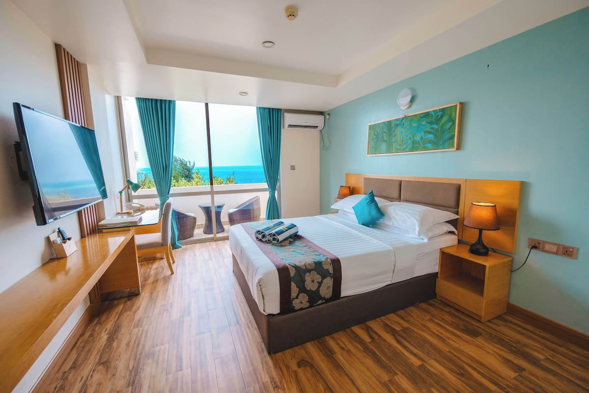 Quarto de hotel com cama, televisão, escrivaninha e vista para o oceano.