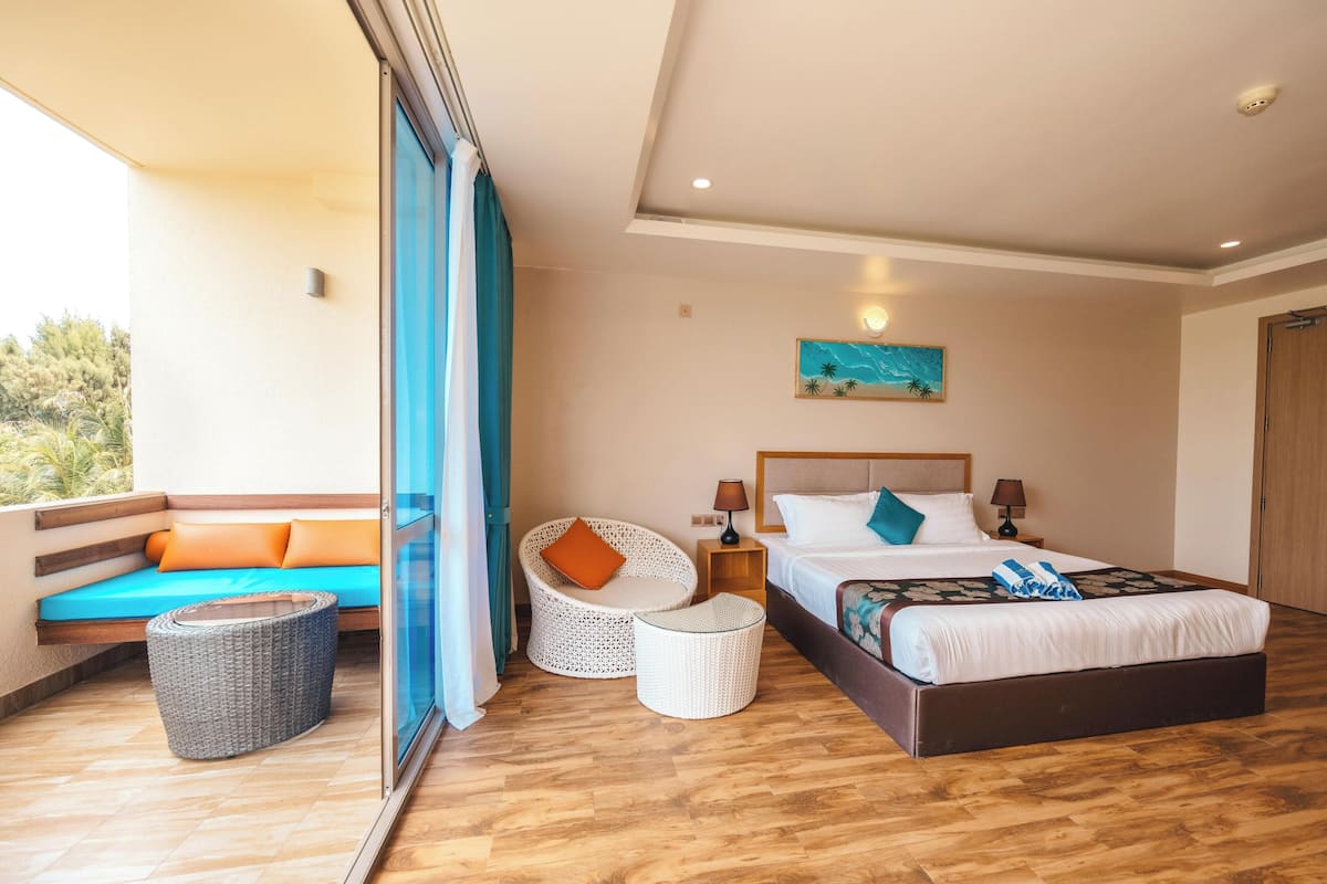 Quarto de hotel com varanda, uma cama com cabeceira de madeira, uma cadeira de vime, um banquinho redondo pequeno e um quadro na parede.