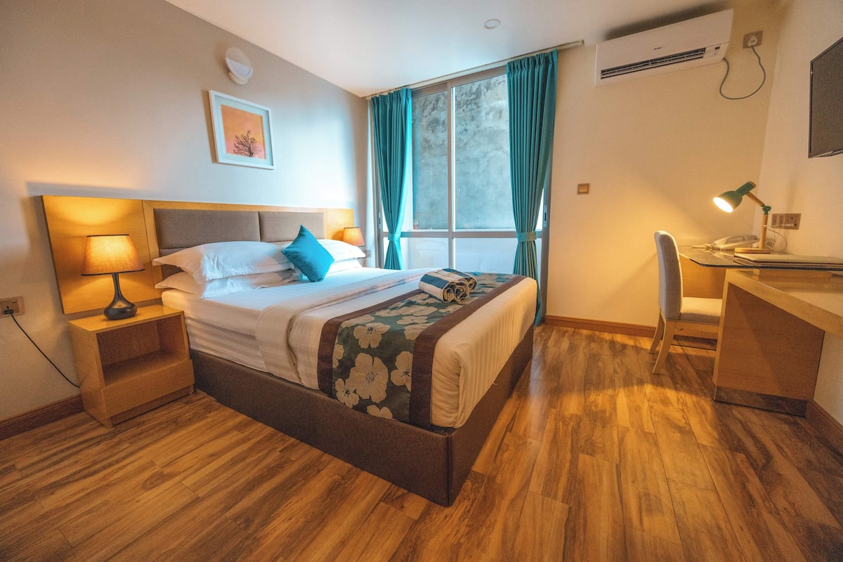 Quarto de hotel com cama, mesa de cabeceira, luminária, escrivaninha, cadeira e janela com cortinas.