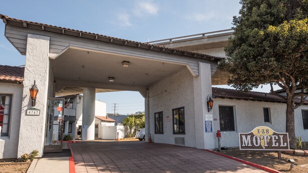 Um motel com entrada coberta, uma placa indicando o nome e o preço, e um estacionamento pavimentado.