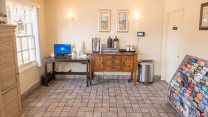 Quarto com escrivaninha de madeira, computador, máquina de café e lixeira. Há duas fotos emolduradas na parede e um mural com revistas.