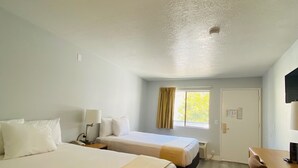 Quarto de hotel com duas camas, uma escrivaninha, uma cadeira, uma janela com cortinas e uma porta.