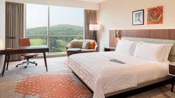 Quarto de hotel com uma cama grande, uma escrivaninha, uma cadeira e vista para uma paisagem verde pela janela.