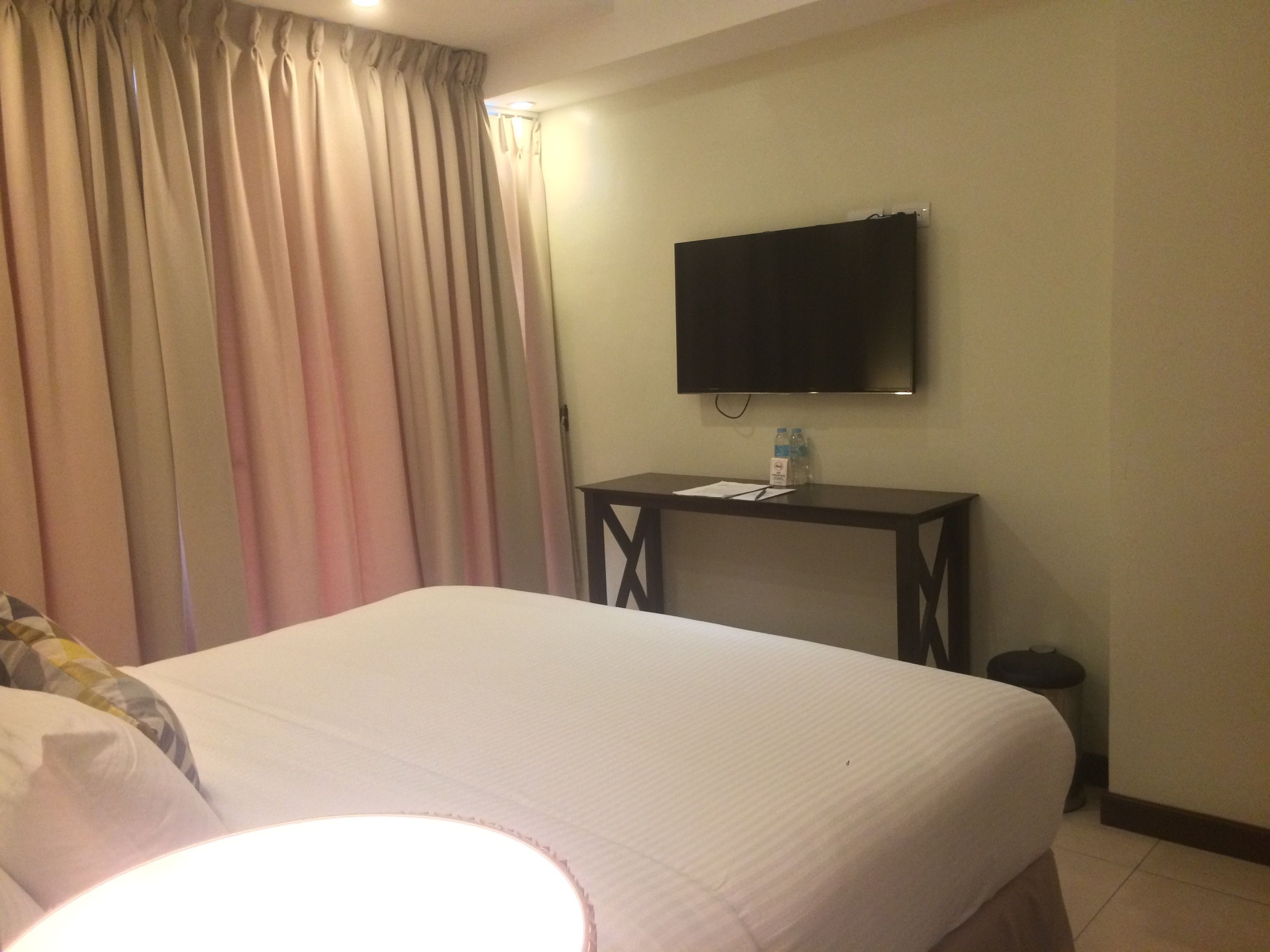Quarto de hotel com uma cama, uma escrivaninha, uma televisão e uma janela com cortinas.
