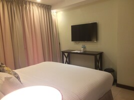 Quarto de hotel com uma cama, uma escrivaninha, uma televisão e uma janela com cortinas.