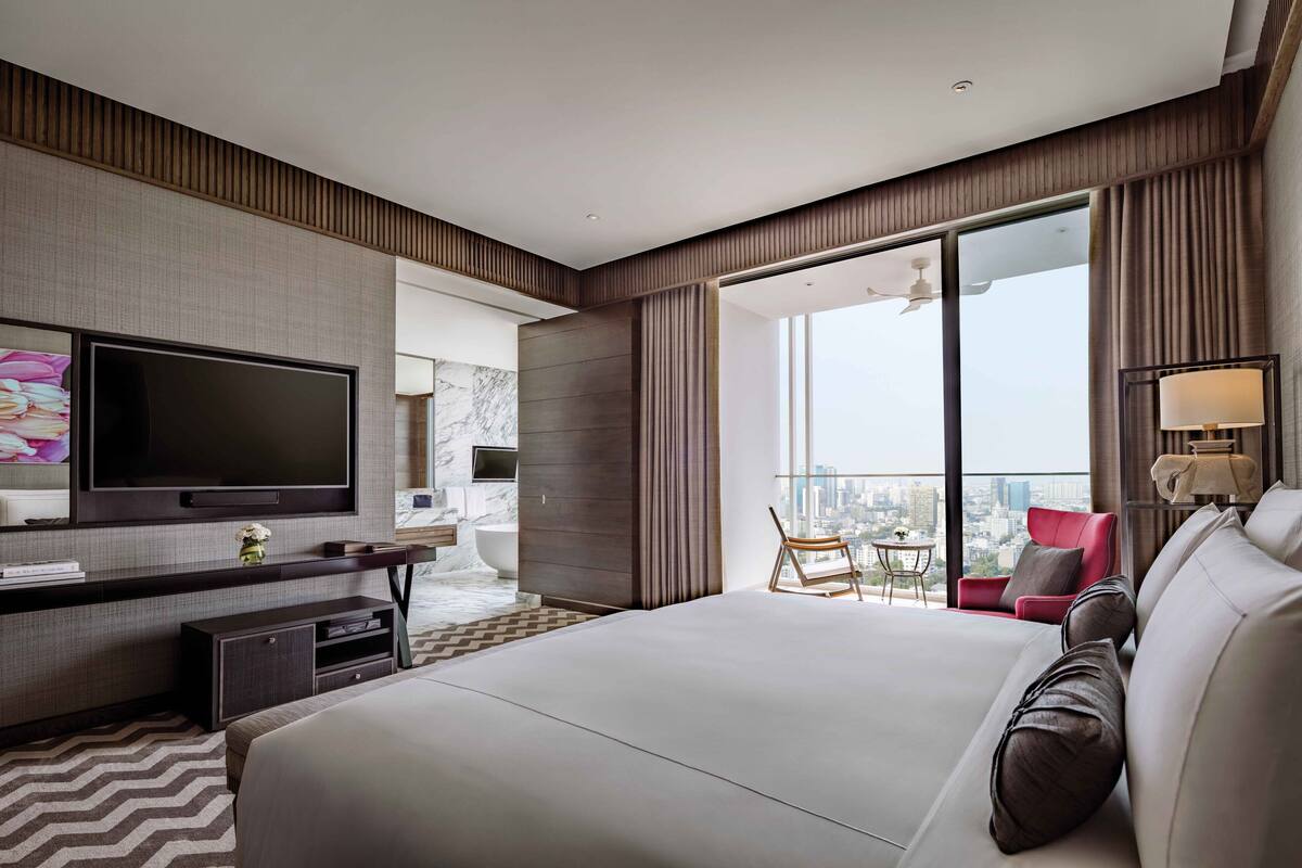 Quarto de hotel moderno com uma cama grande, televisão de tela plana, varanda com vista para a cidade e banheiro separado.