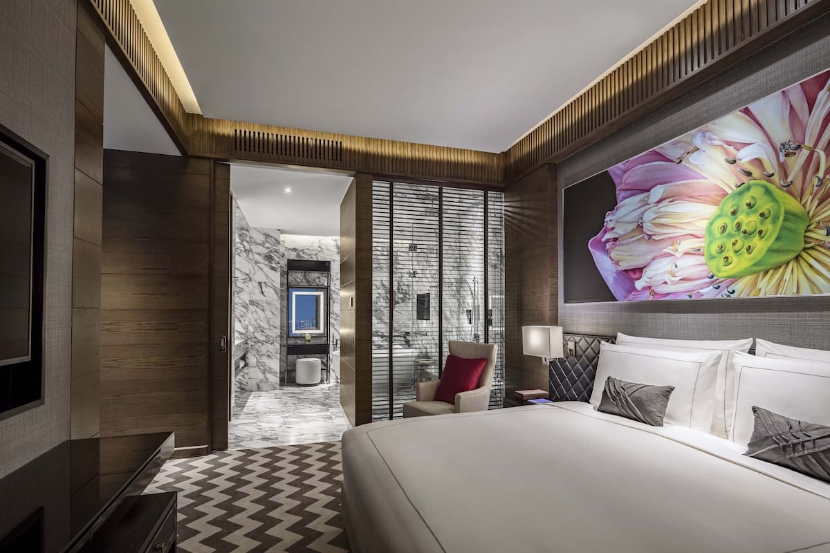 Quarto de hotel moderno com uma cama grande, televisão e casa de banho visível através de uma porta aberta.