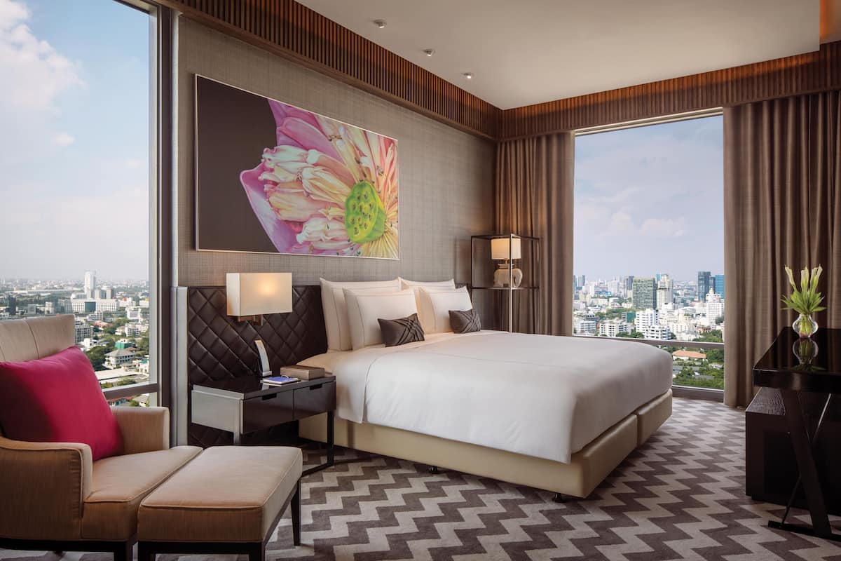 Quarto de hotel moderno com uma cama grande, vista para a cidade, um quadro floral grande na parede e uma escrivaninha com luminária.