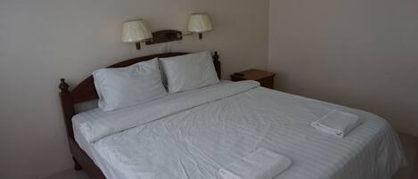 Cama bem arrumada com roupa de cama e toalhas brancas, cabeceira de madeira, duas luminárias de mesa e uma mesinha de cabeceira.