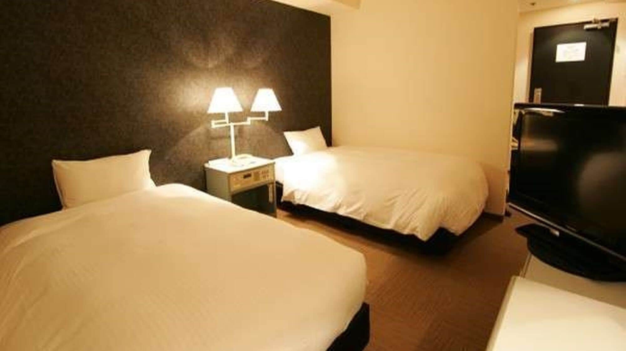 Quarto de hotel com duas camas, uma escrivaninha com abajur, uma televisão e uma porta.