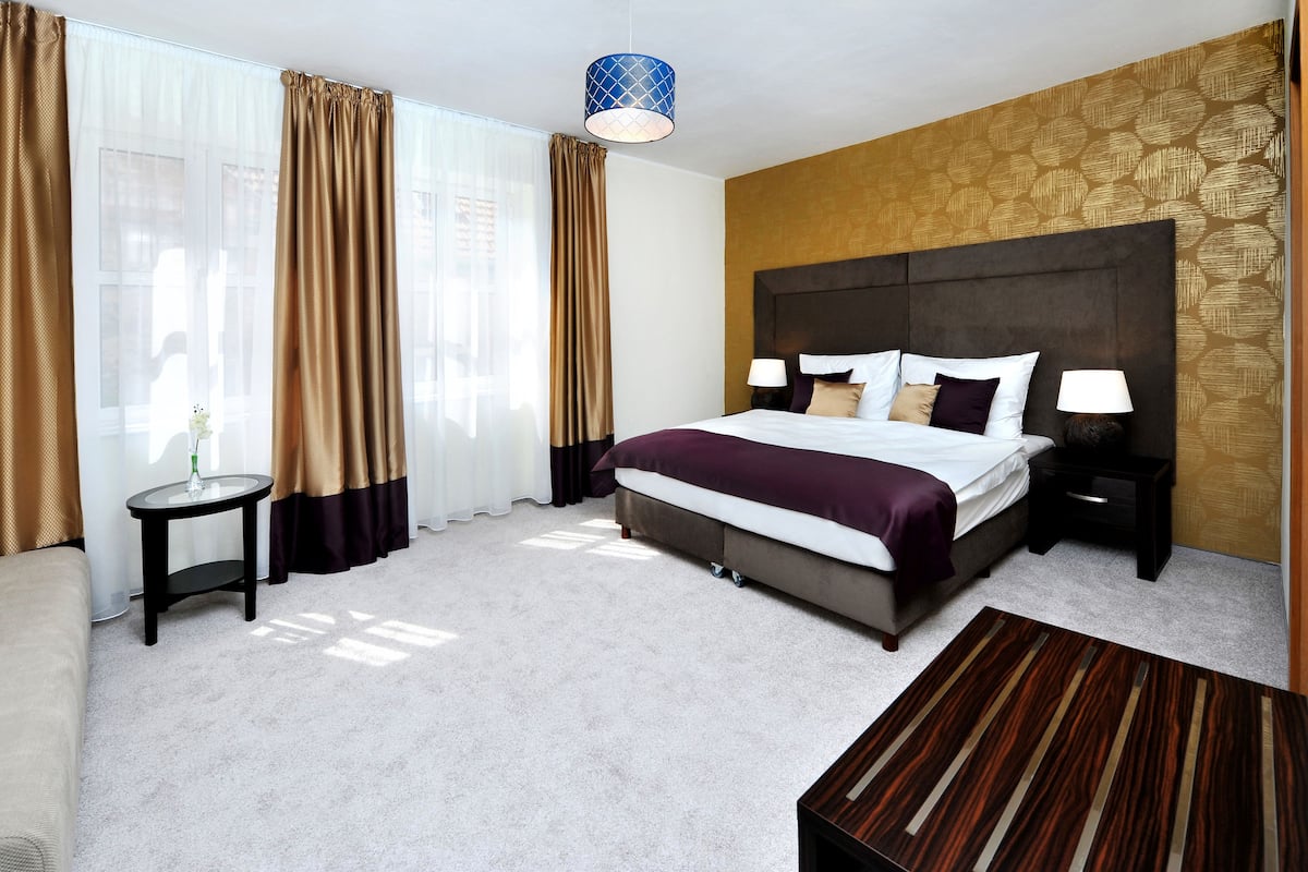 Quarto de hotel com uma cama grande, duas mesinhas de cabeceira, uma mesa lateral redonda, uma mesa de centro de madeira e uma parede estampada.