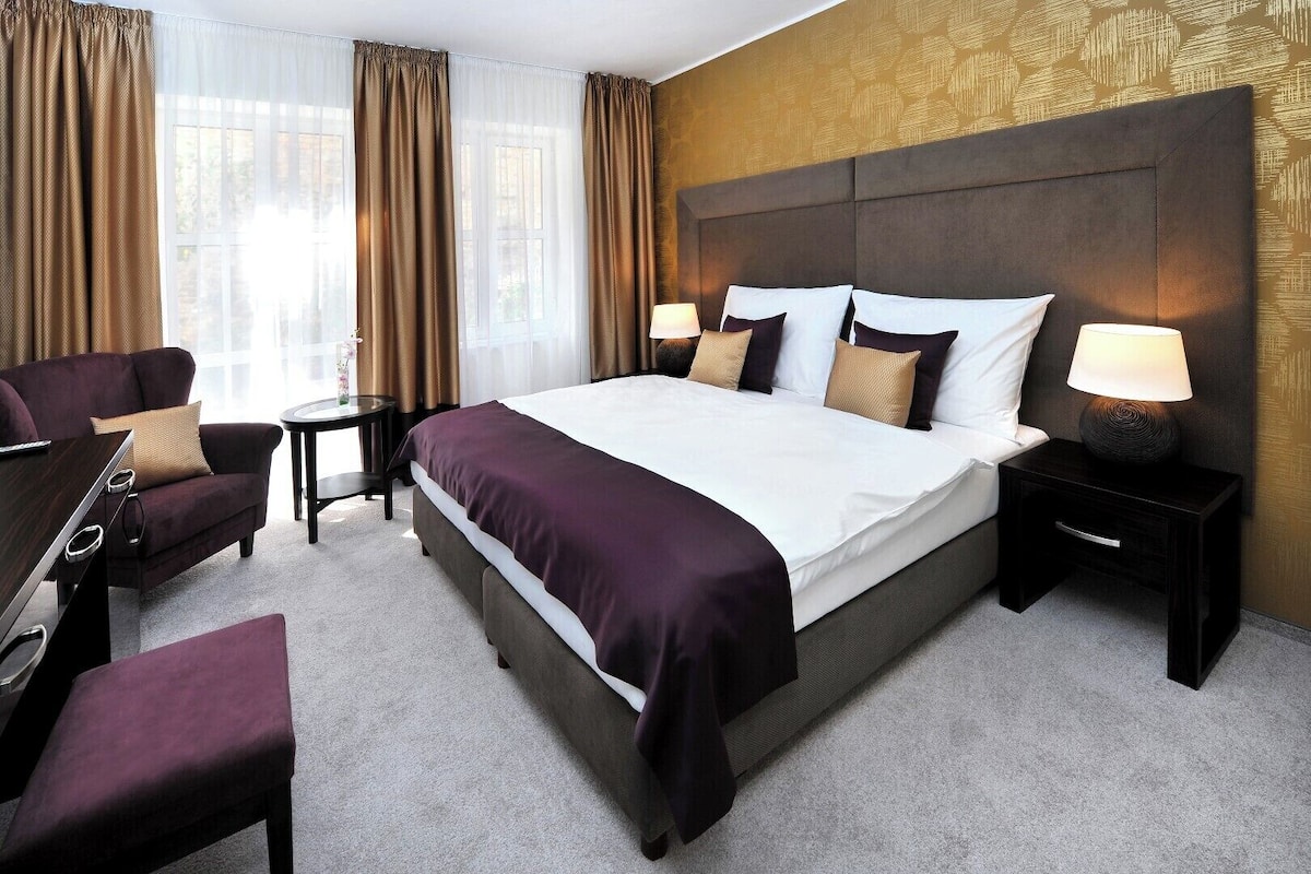 Quarto de hotel com uma cama grande, uma escrivaninha, uma cadeira e uma mesa de cabeceira com abajur.