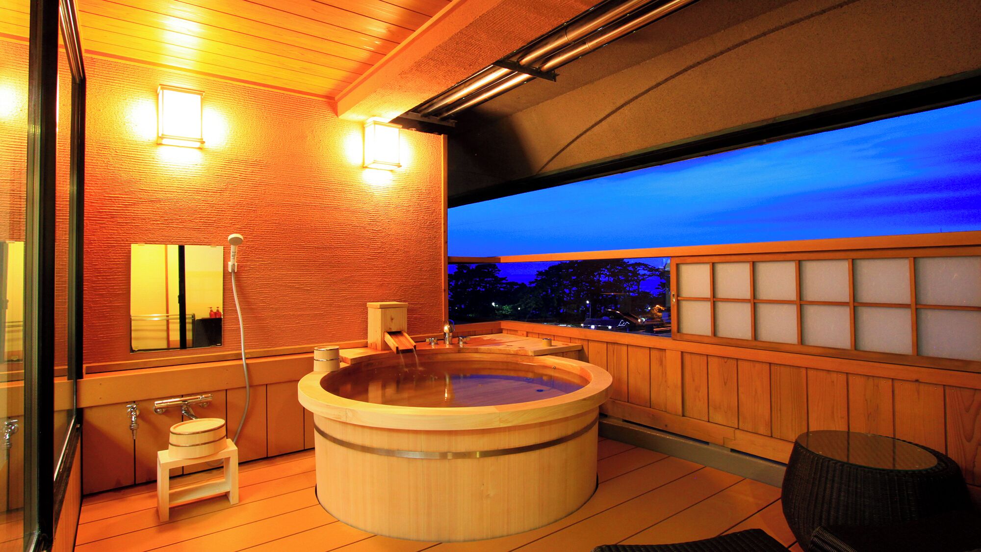 Um spa de madeira com hidromassagem, um banquinho de madeira pequeno e vista para a cidade ao anoitecer.
