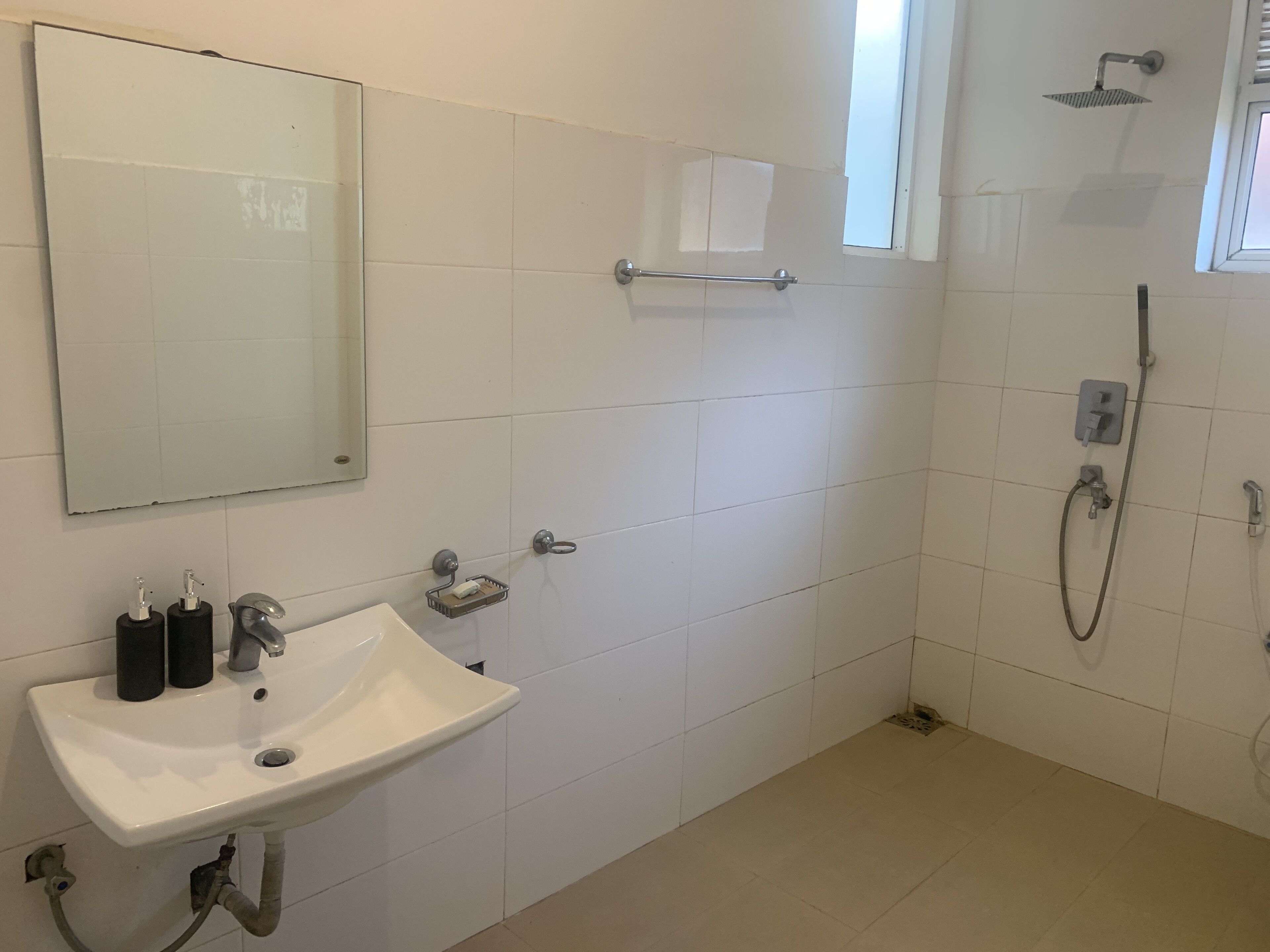 Casa de banho com lavatório branco, espelho, porta-toalhas, chuveiro com chuveiro manual e janela.
