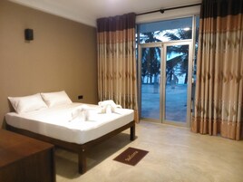 Quarto de hotel com uma cama, mesa de cabeceira, janela com cortinas e vista para palmeiras e uma praia.
