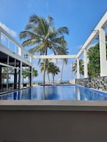Área de piscina com vista para o oceano, palmeiras e uma construção moderna.