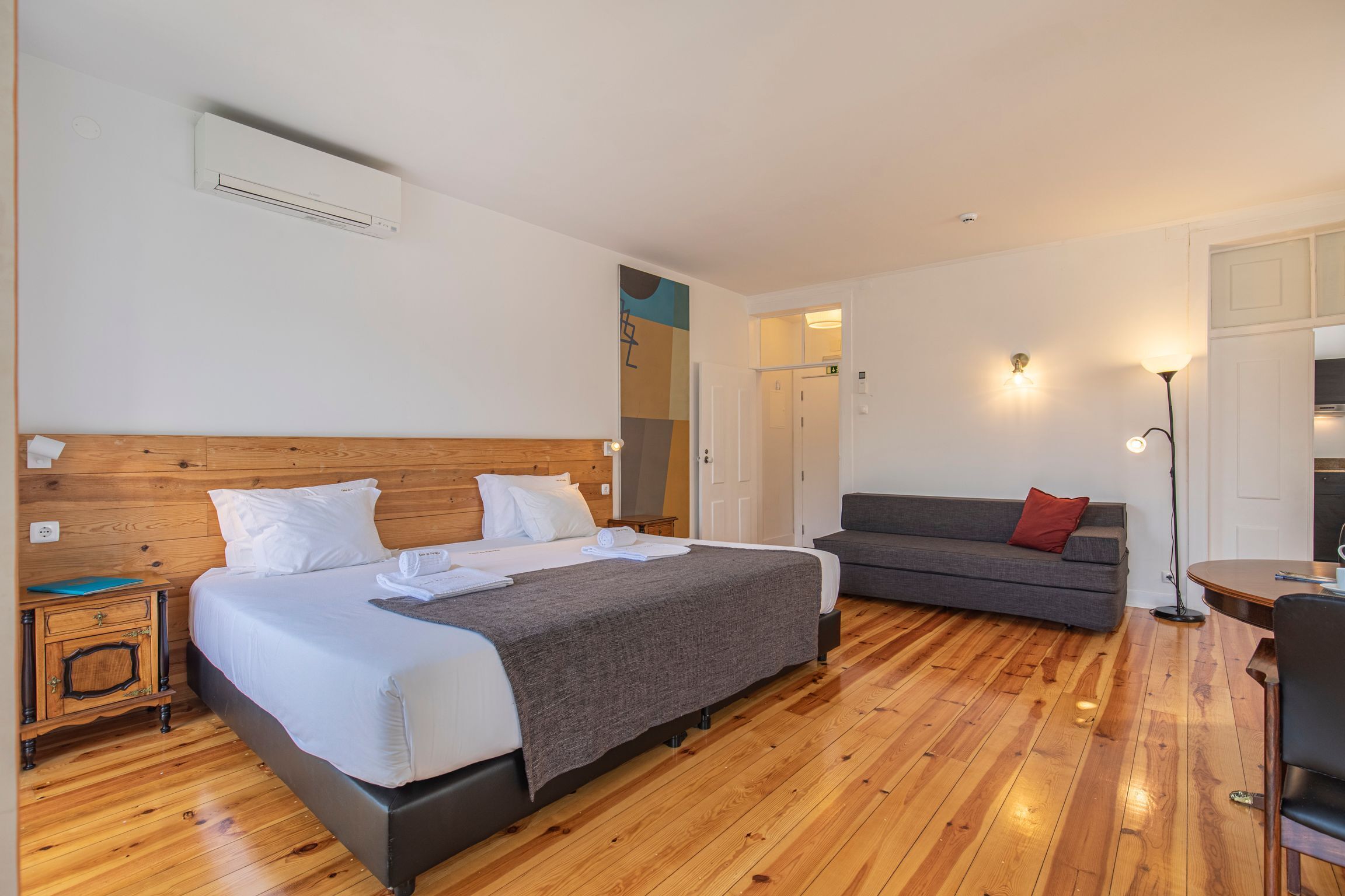 Quarto de hotel com piso de madeira, uma cama de casal com roupa de cama branca, um sofá cinza, uma mesa pequena e um ar condicionado fixado na parede.
