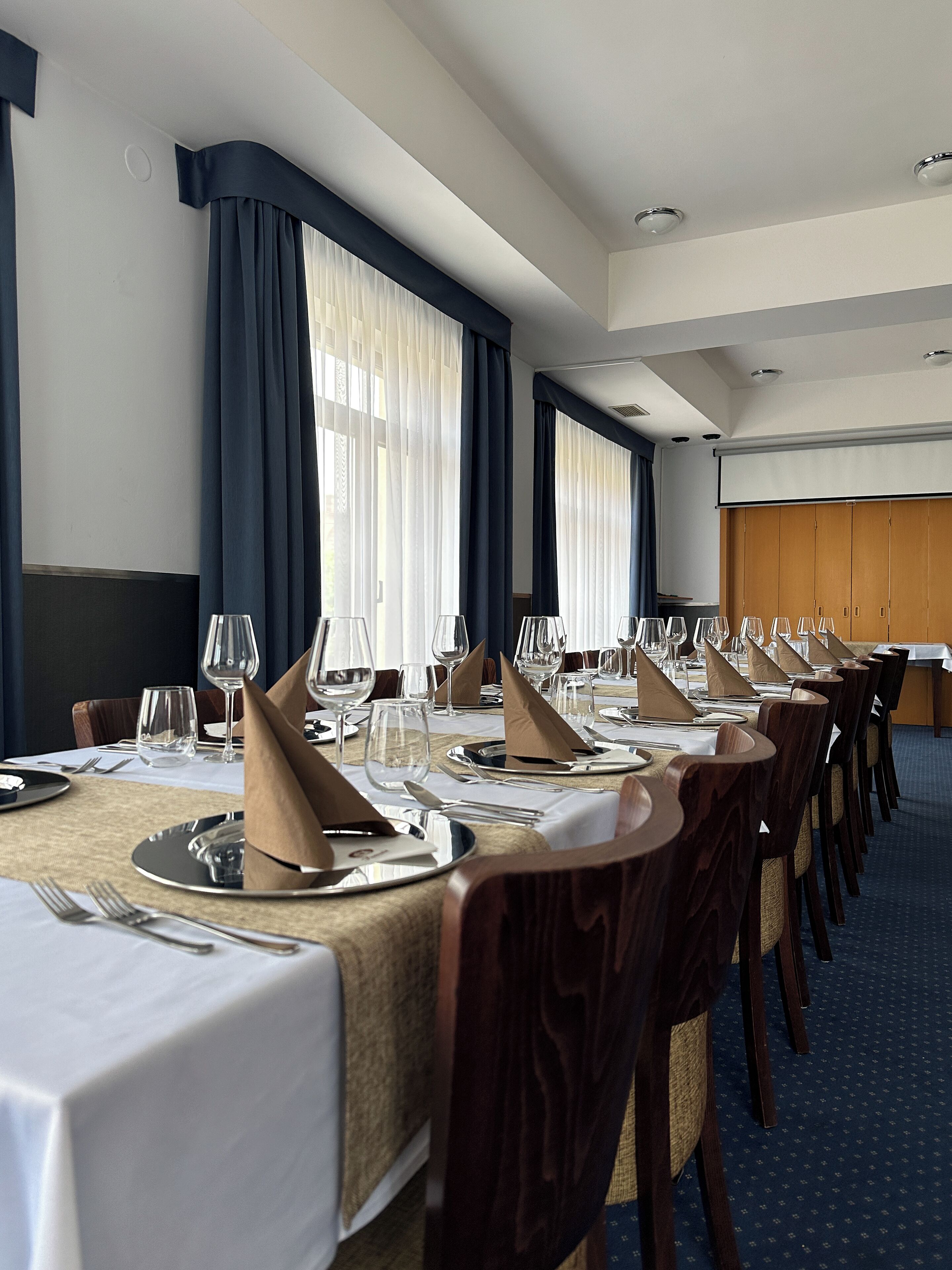 Sala de conferências com mesas compridas preparadas para uma reunião, janelas amplas com cortinas e uma tela de projeção na parede.