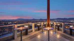 Terraço no telhado com mesas e cadeiras, oferecendo uma vista panorâmica da cidade ao entardecer.