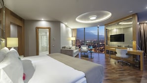 Quarto de hotel com cama grande, escrivaninha, cadeira, sofá e televisão. Oferece vista para a paisagem urbana através de grandes janelas.