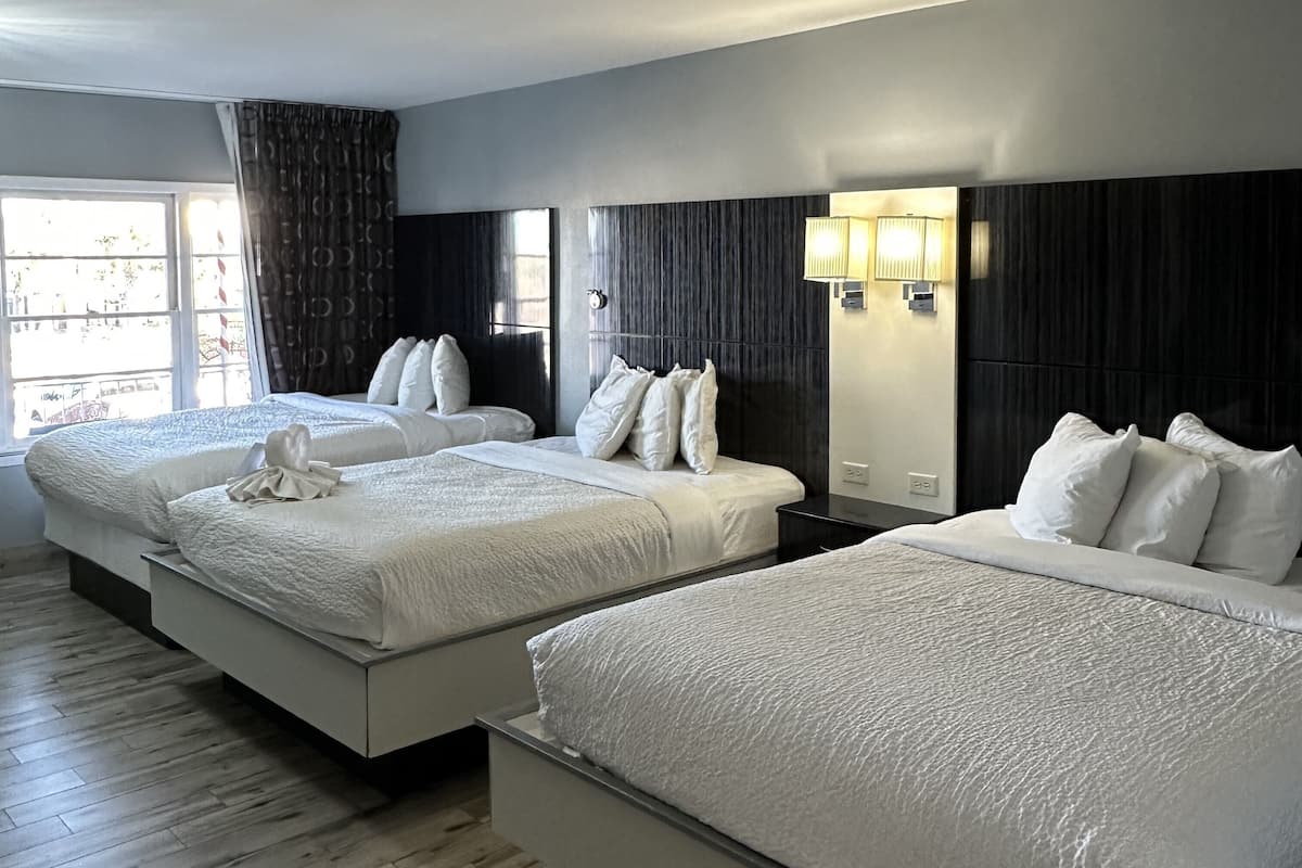 Quarto de hotel com duas camas, uma janela grande e uma cabeceira escura com luzes acopladas à parede.