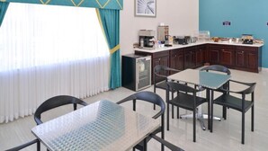 Uma área de refeições com mesa e cadeiras, um espaço para café com máquina e xícaras, e um balcão com geladeira.