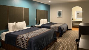 Quarto de hotel com duas camas, uma escrivaninha e um banheiro visível através de um arco.