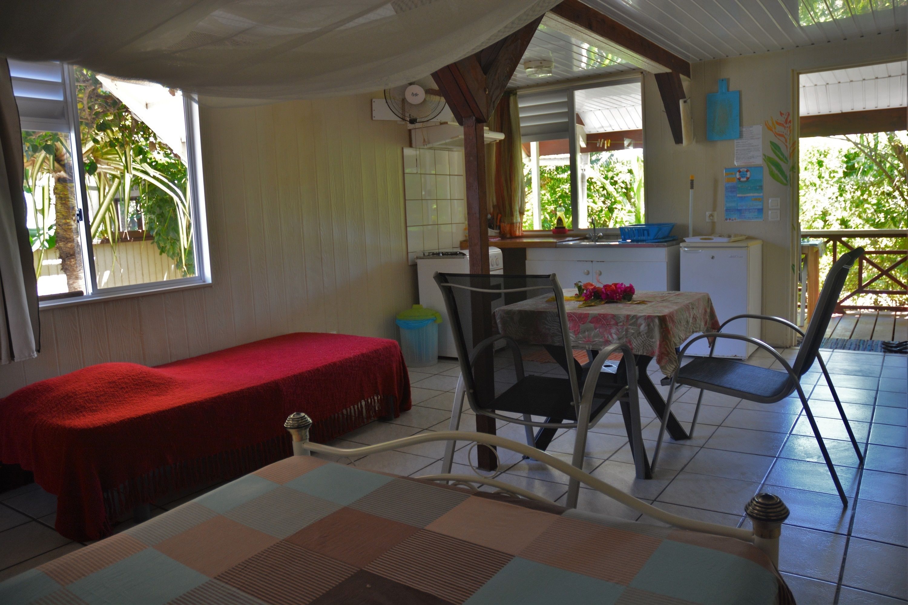 Quarto com cama, mesa e cadeiras, pequena cozinha e vista para o verde lá fora.