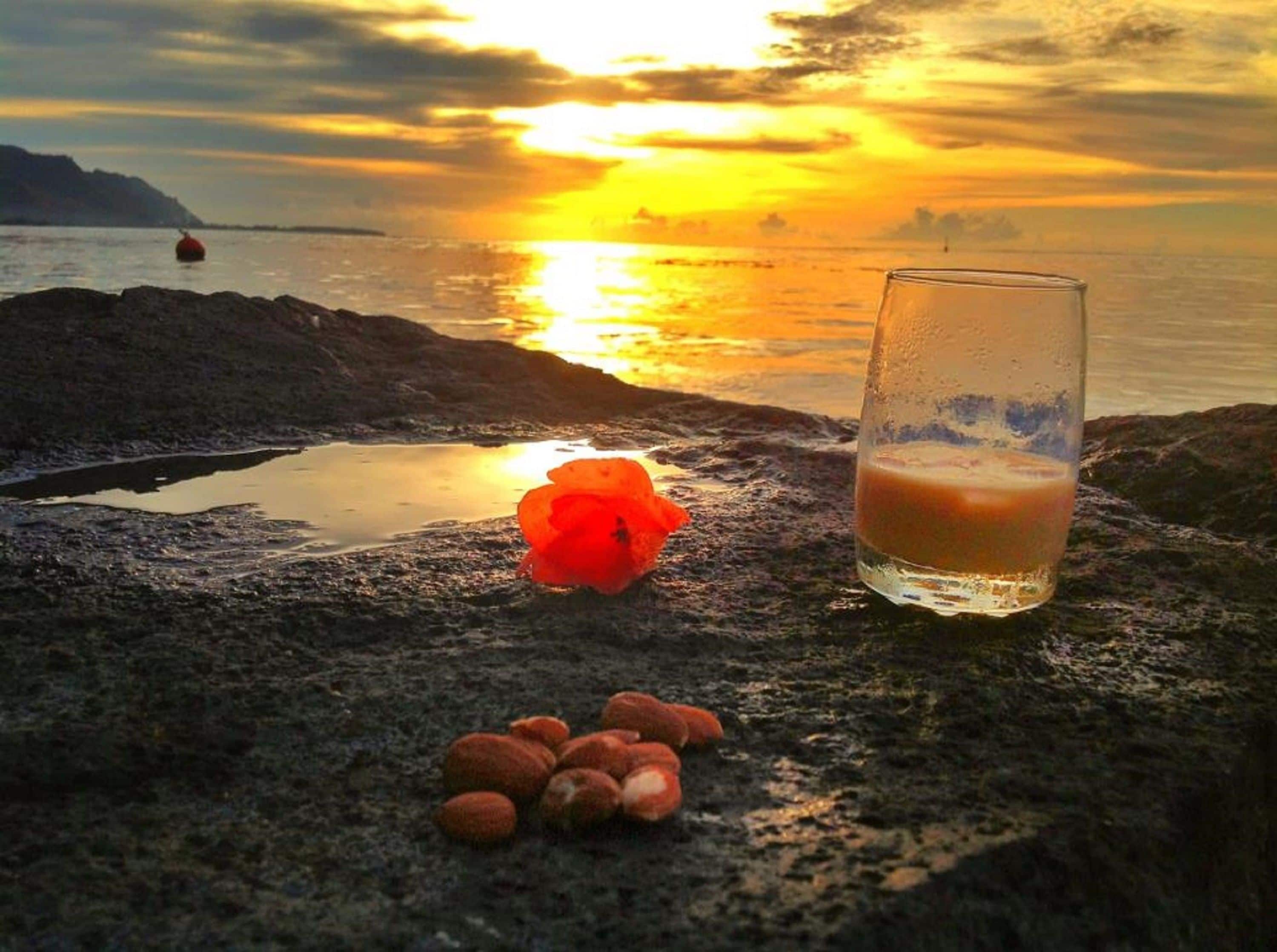 Um copo com líquido laranja sobre uma superfície rochosa, com um pôr do sol ao fundo.