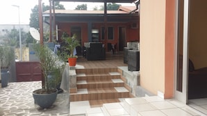 Área externa coberta com escadaria para a entrada de um edifício, plantas em vasos e mobiliário de exterior.