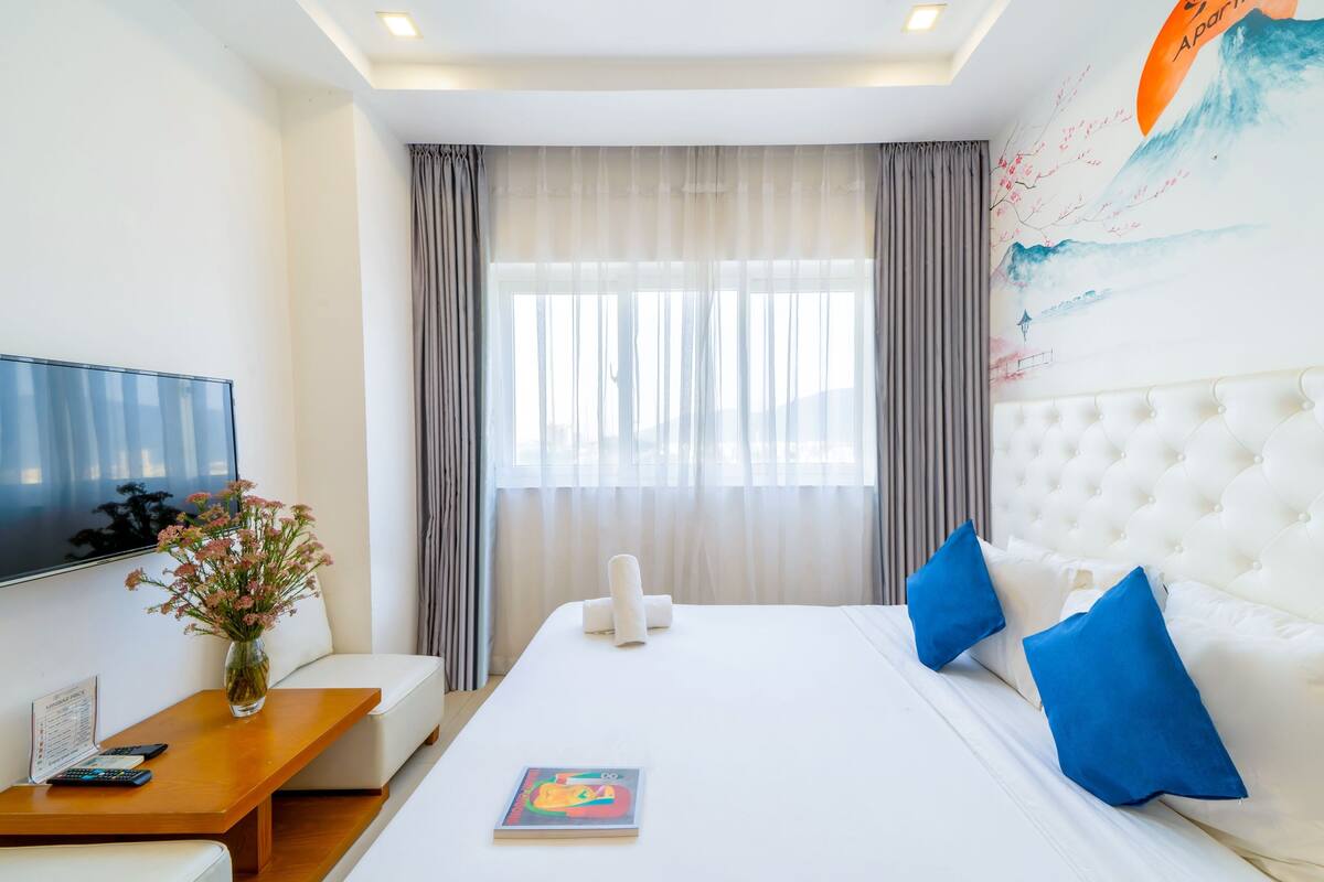 Quarto de hotel com uma cama grande, televisão de tela plana, mesa de cabeceira de madeira e um vaso com flores.