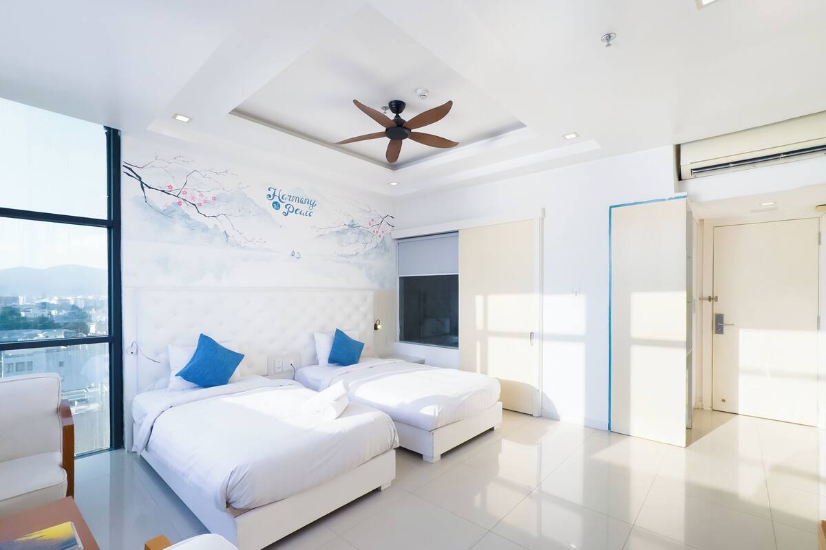 Quarto de hotel moderno com duas camas, um ventilador de teto, uma janela ampla com vista para a cidade e uma porta à direita.