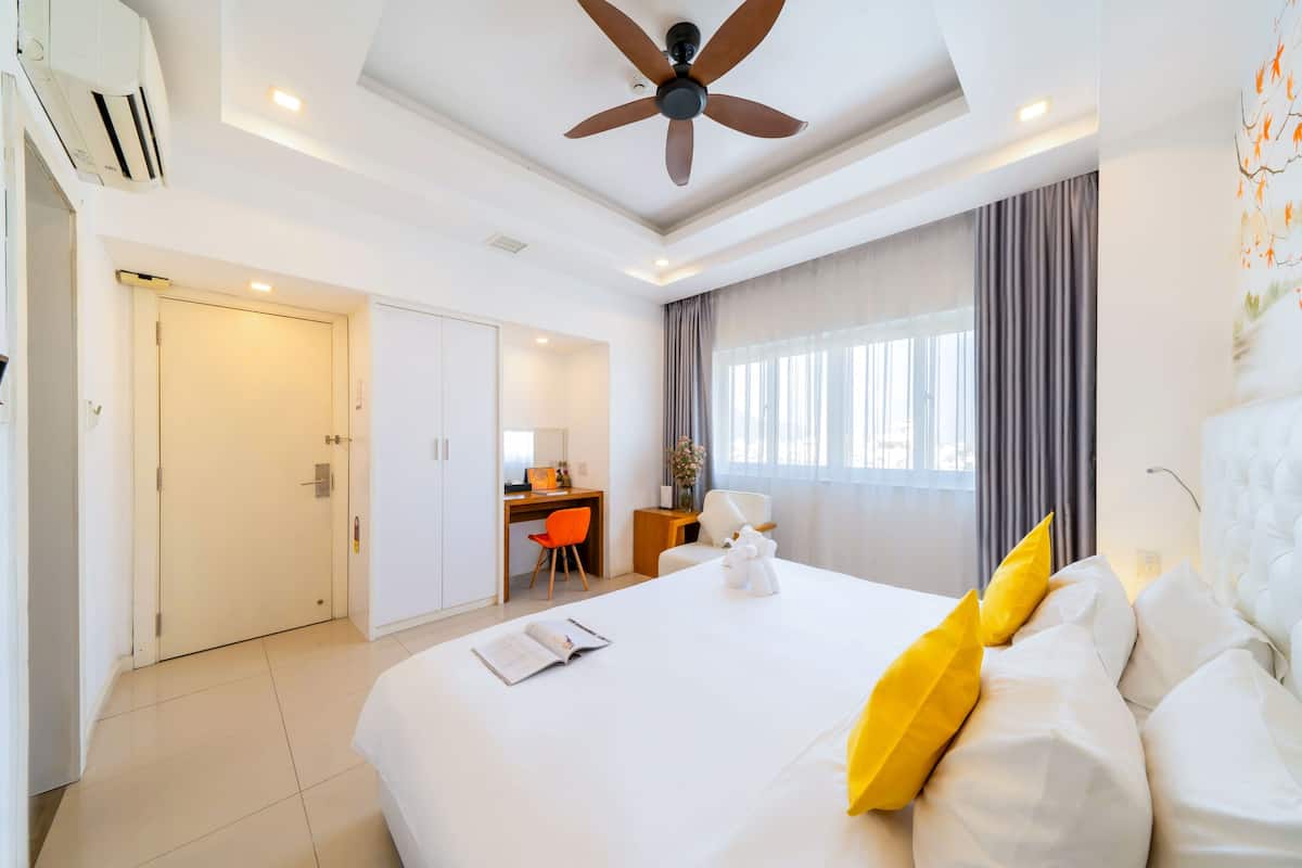 Quarto de hotel com cama, escrivaninha, cadeira, ventoinha no teto e janela com cortinas.