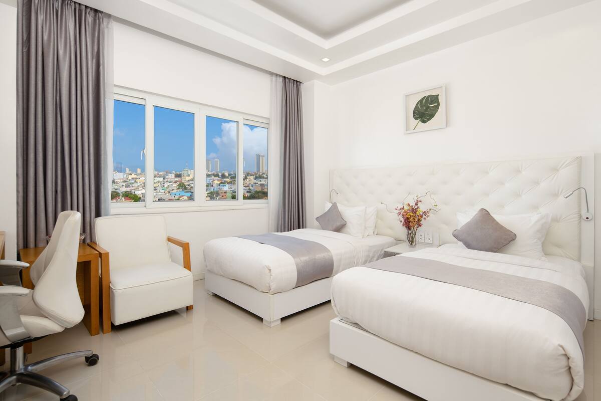 Quarto de hotel com duas camas, uma mesa, uma cadeira e vista para a paisagem urbana através de janelas amplas.