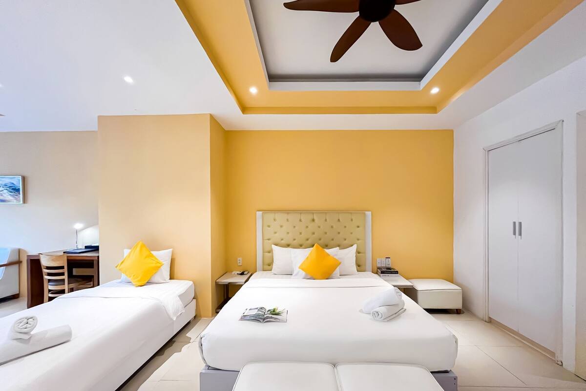 Quarto de hotel com duas camas, uma escrivaninha, uma cadeira, um ventilador de teto e um guarda-roupa.