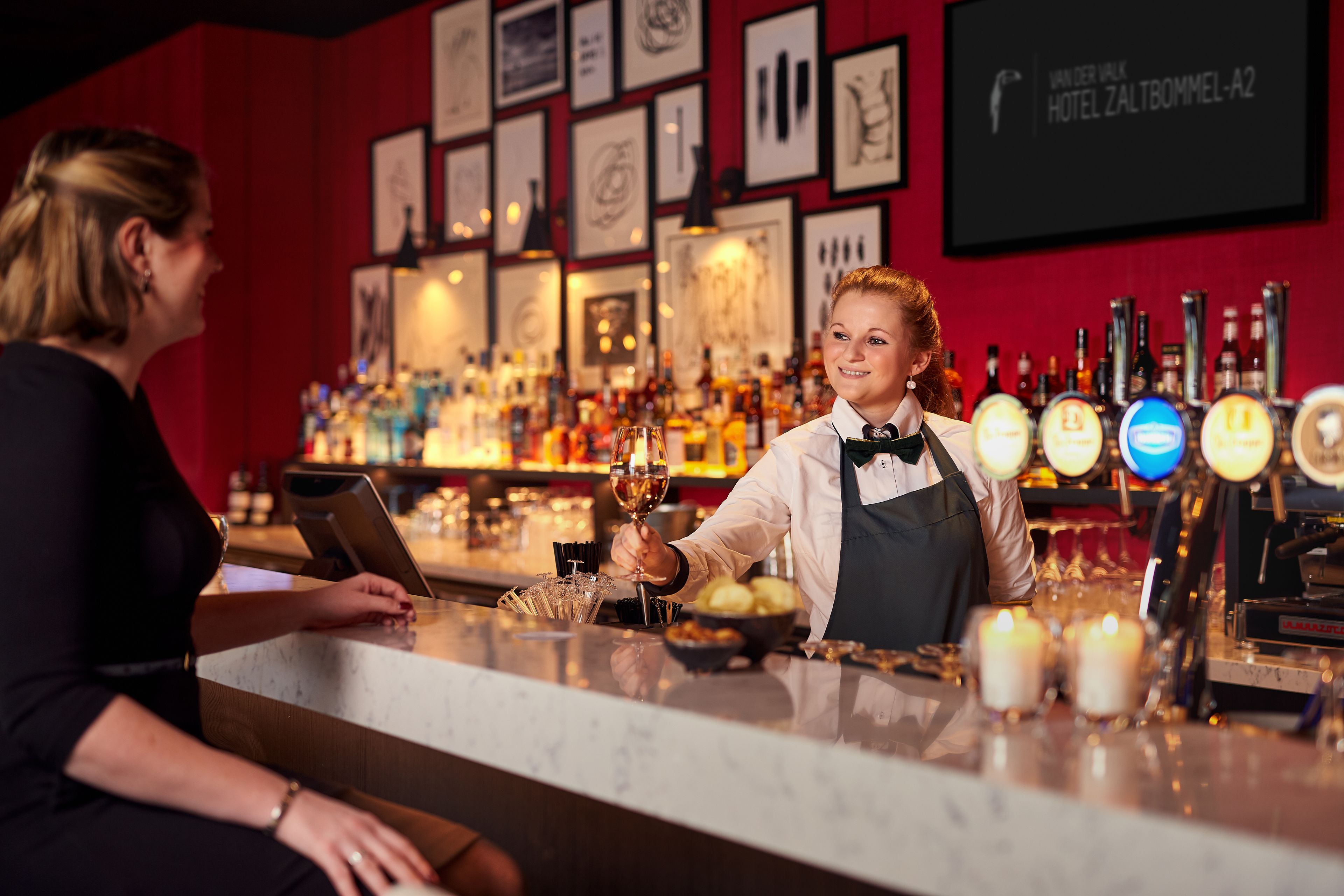 Uma área de bar com um bartender, um cliente e um bar bem abastecido com diversos garrafas e copos.