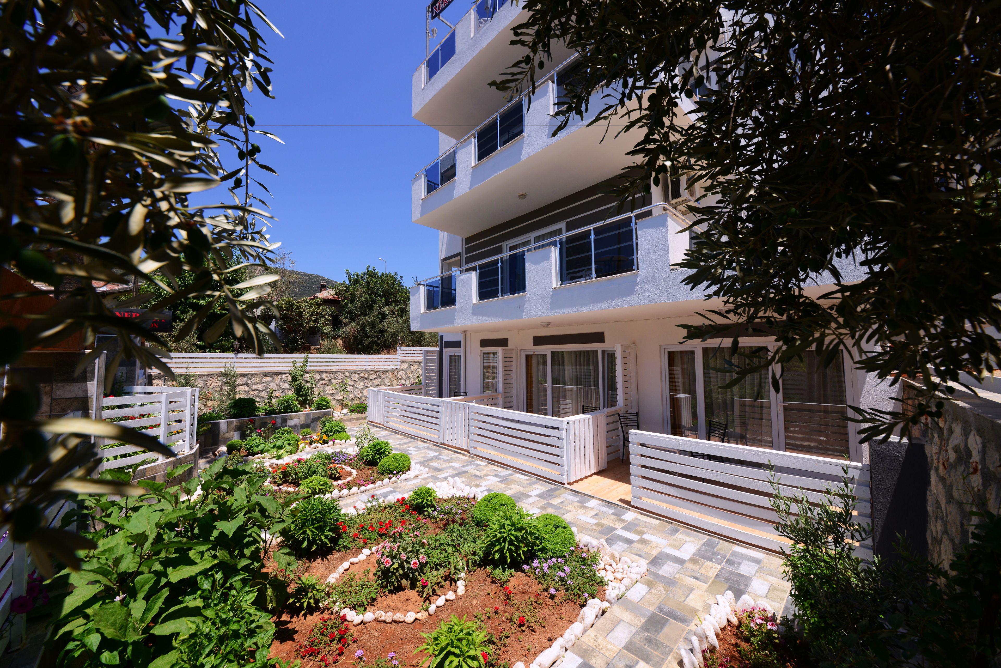 Um edifício moderno de vários andares com varandas, um jardim com diversas plantas e um pavimento xadrez.
