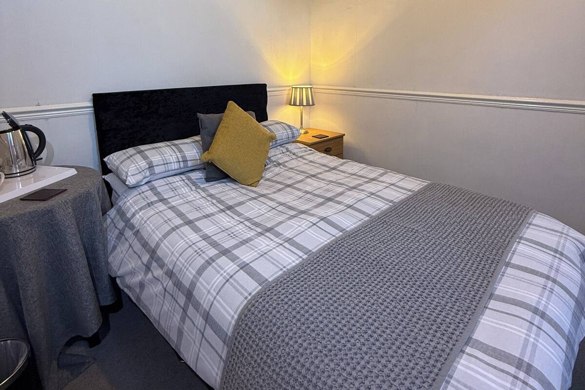 Cama bem arrumada com colcha xadrez e um único travesseiro amarelo. Tem uma mesinha de cabeceira com abajur e chaleira sobre ela. O quarto tem paredes lisas e um design simples e limpo.