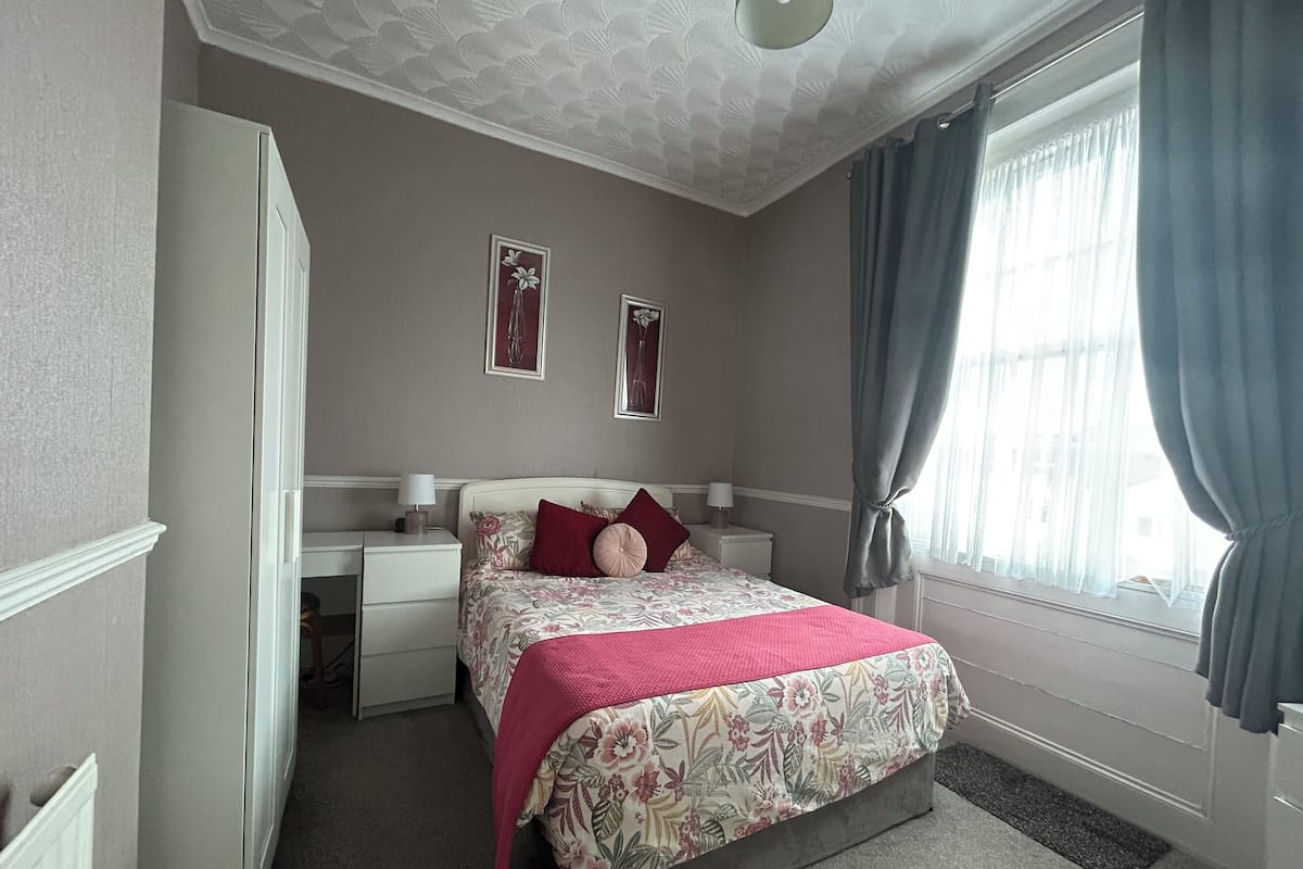 Quarto com cama, mesas de cabeceira, cômoda, janela com cortinas e duas imagens penduradas na parede.