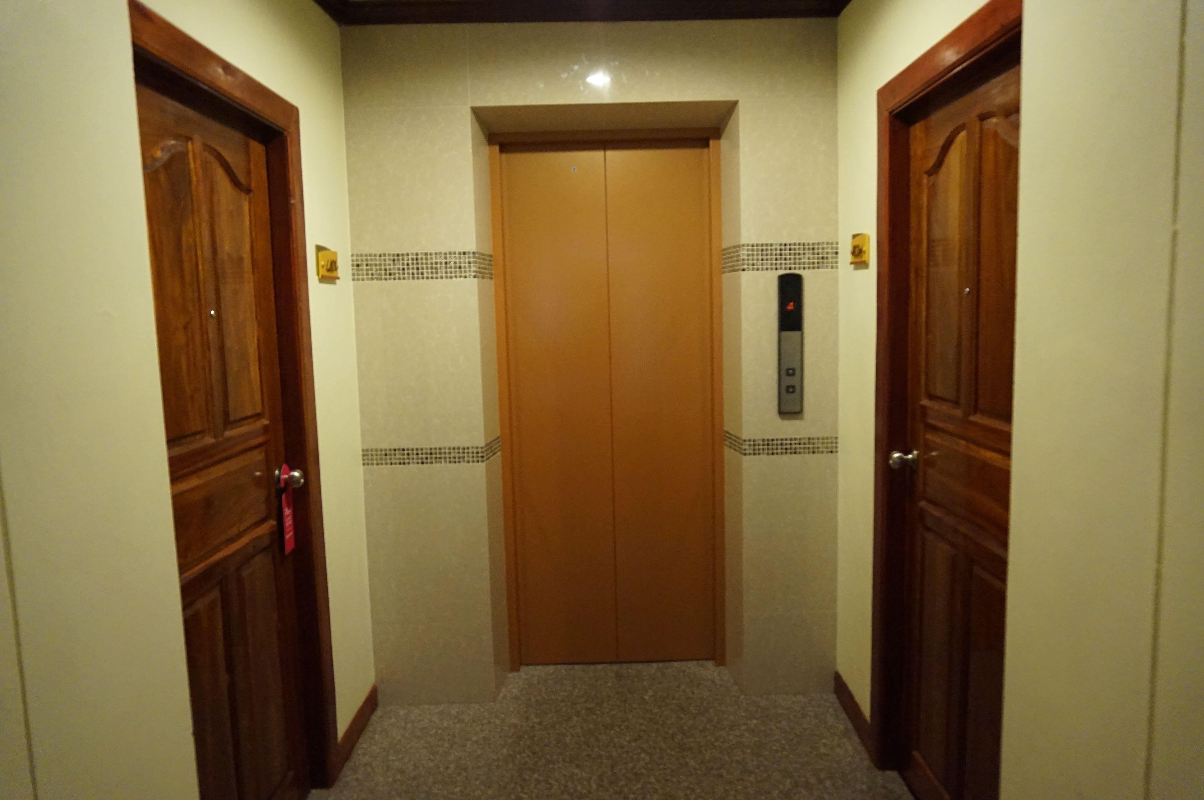 Um corredor com uma porta de madeira no final, um elevador fixado na parede e uma porta com etiqueta vermelha.